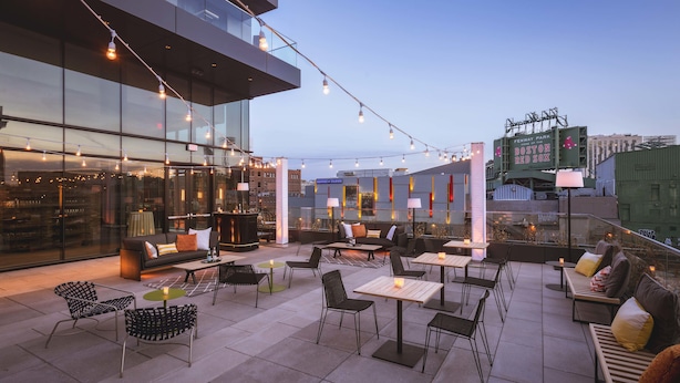 Uma área externa com mesas, cadeiras, um bar e vista para o Fenway Park.