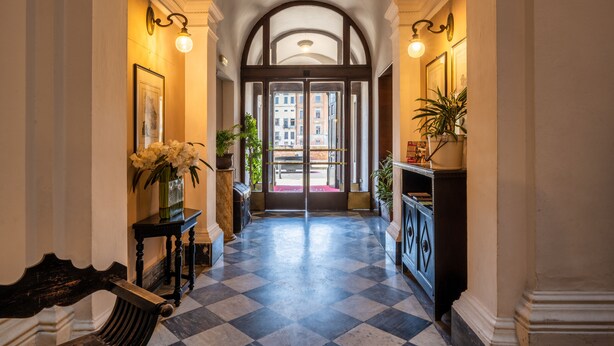 Um saguão de hotel com piso xadrez, uma escadaria imponente e vista da cidade pelas portas.
