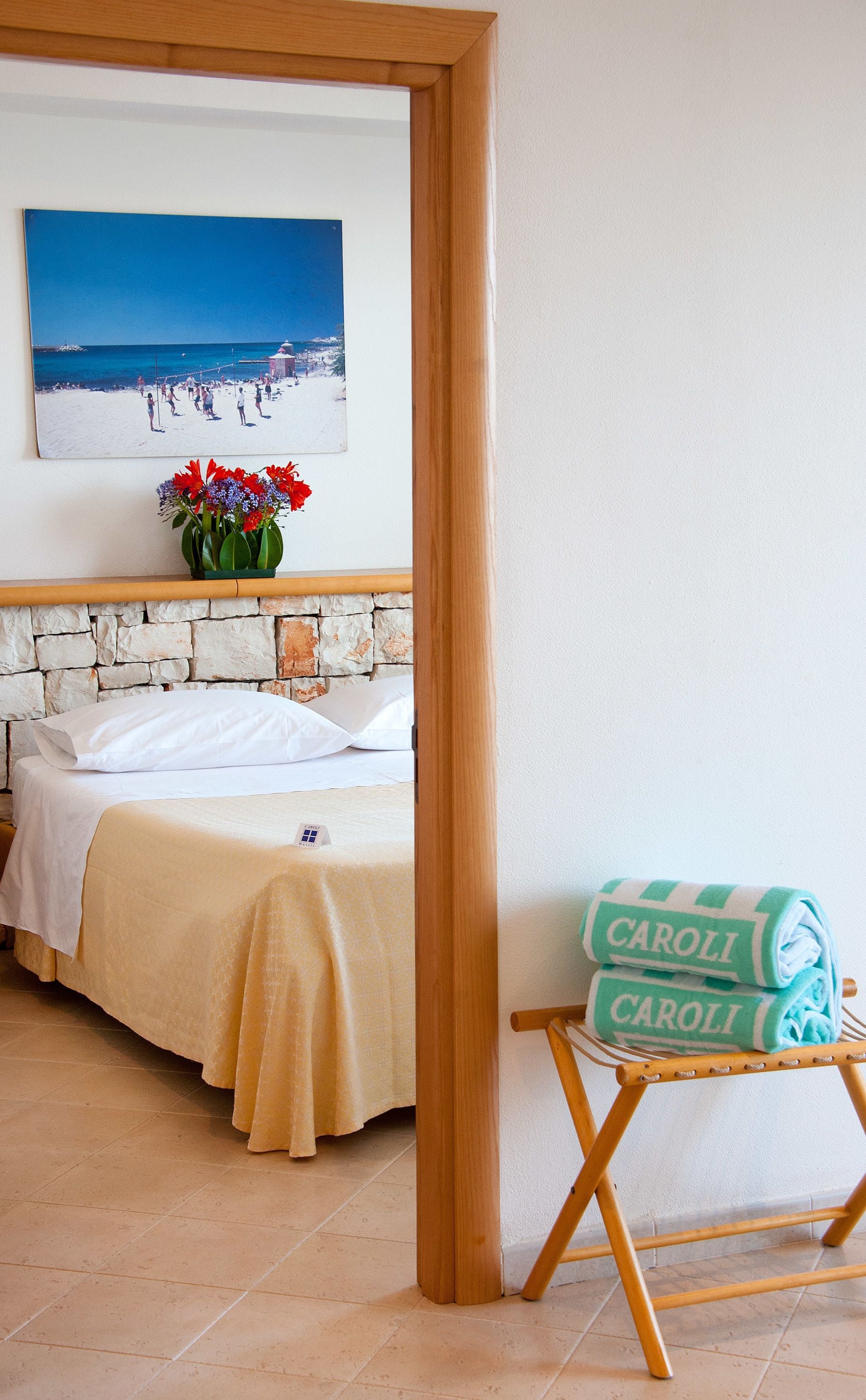 Quarto com parede de pedra, cama com lençóis brancos, pintura de uma cena de praia e uma pequena mesa com toalhas.