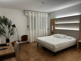Quarto de hotel moderno com soalho de madeira, cama com roupa de cama branca, secretária com cadeira, planta em vaso e uma janela grande com cortinas brancas.