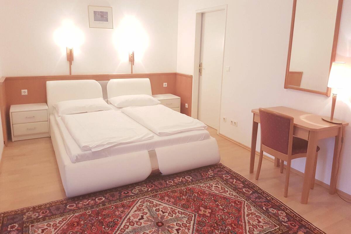 Quarto de hotel com cama de casal, mesas de cabeceira, escrivaninha com cadeira, espelho e tapete estampado.