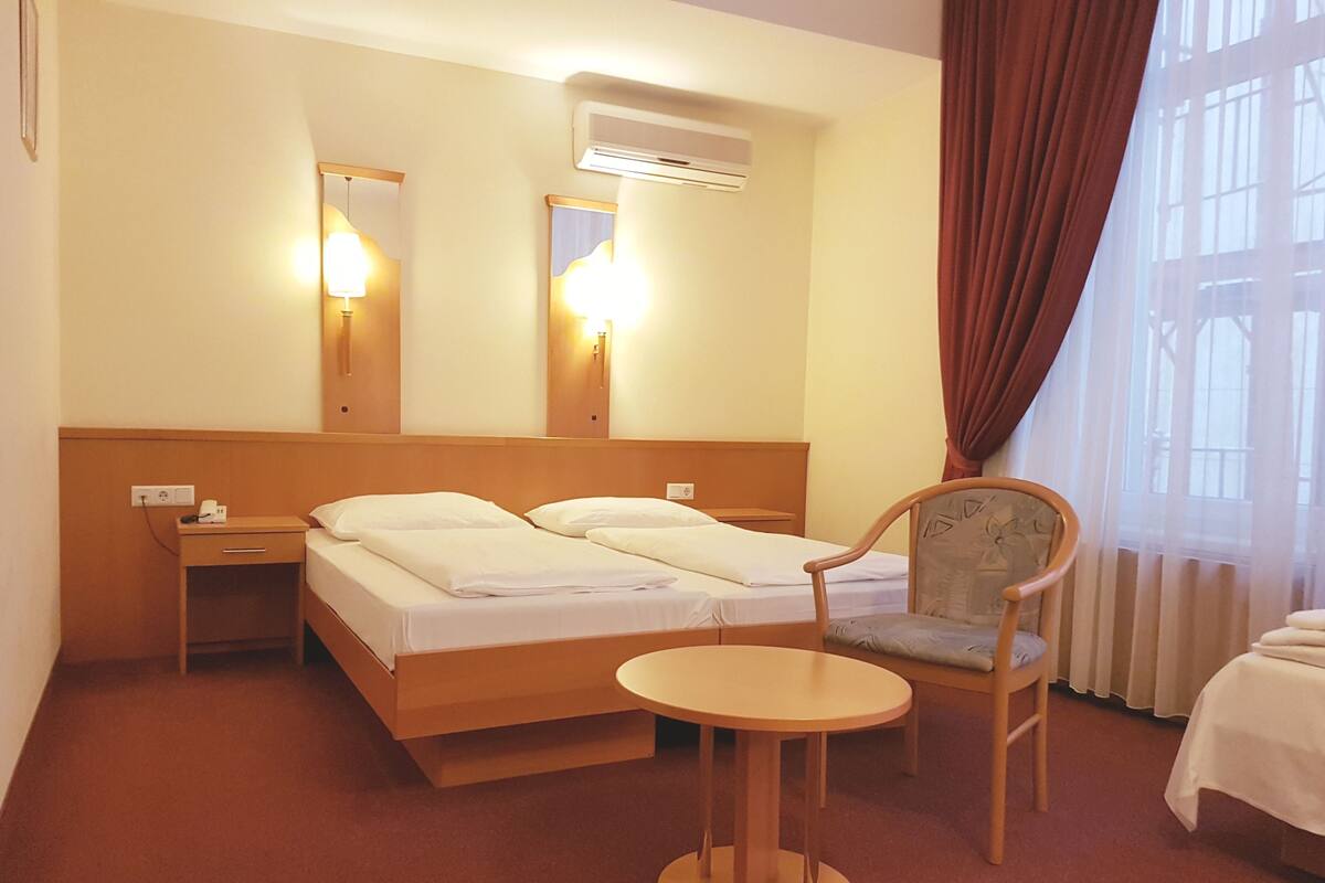 Quarto de hotel com cama, mesas de cabeceira, uma cadeira, uma mesinha redonda, ar condicionado e uma janela com cortinas.