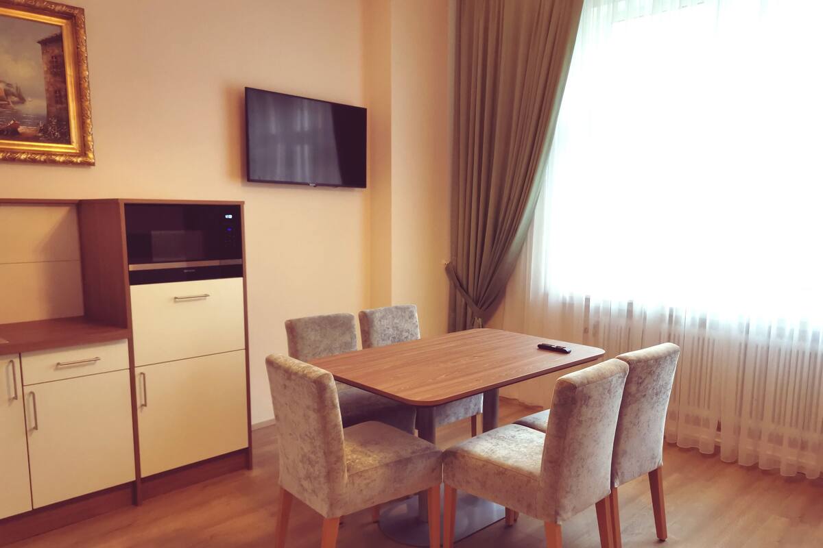 Uma sala de jantar com mesa de madeira e quatro cadeiras, televisão fixada na parede e uma janela com cortinas.