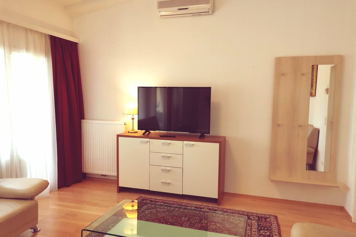 Uma sala de estar com televisão de tela plana em um móvel de madeira, mesa de centro de vidro, tapete vermelho estampado e sofá bege.