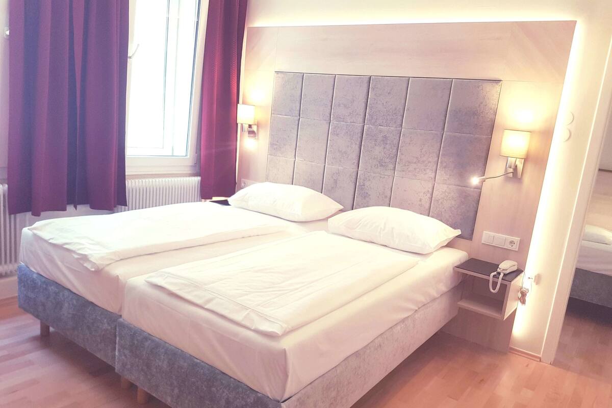 Uma cama de casal com cabeceira cinza, roupa de cama branca e duas luminárias de apoio.