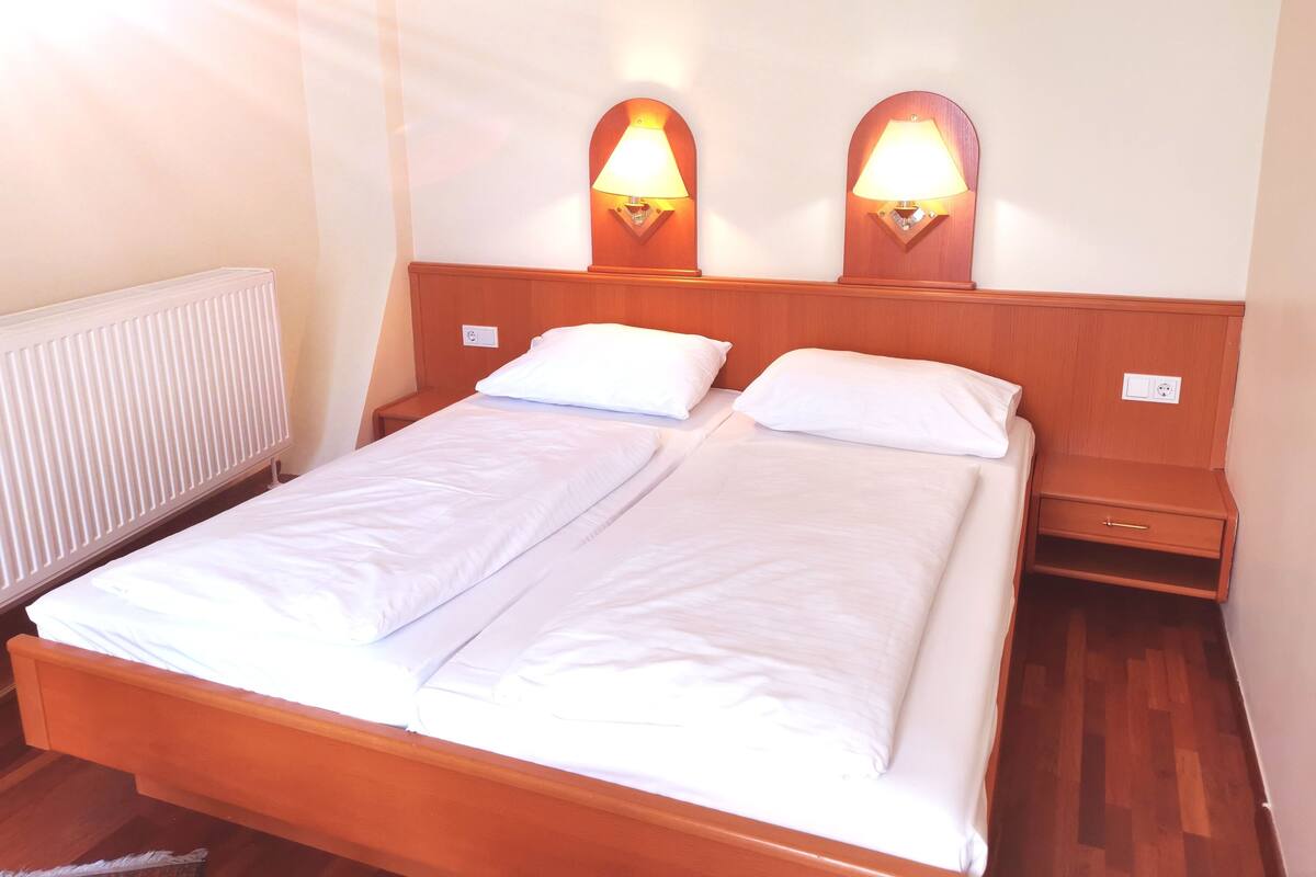 Uma cama de casal com lençóis brancos, duas mesas de cabeceira com abajures e uma cabeceira de madeira.