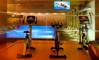 Uma academia com bicicletas ergométricas, esteira e piscina ao fundo.