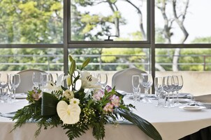 Mesa posta para refeição, com arranjo floral, copos de vidro transparentes e vista para árvores através de grandes janelas.