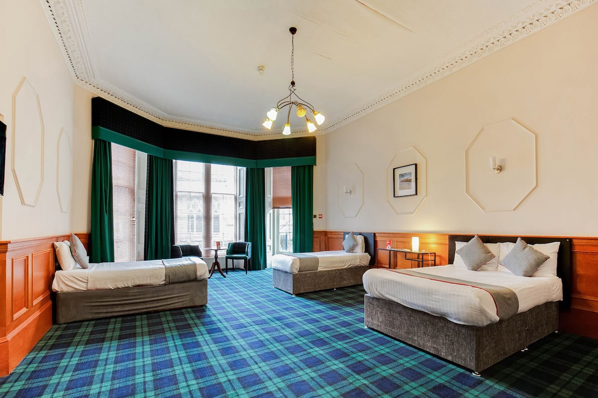 Quarto de hotel com duas camas, uma janela grande com cortinas, piso xadrez e um lustre.