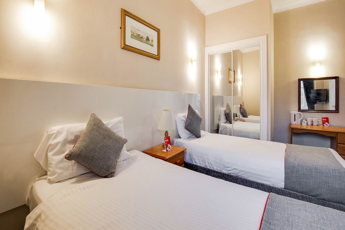 Quarto de hotel com duas camas, uma mesinha de cabeceira com abajur, um espelho e um quadro na parede.