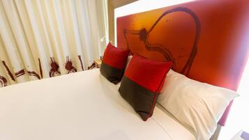 Quarto de hotel com uma cama grande, duas almofadas vermelhas e uma branca, cabeceira de madeira e uma janela com persianas.