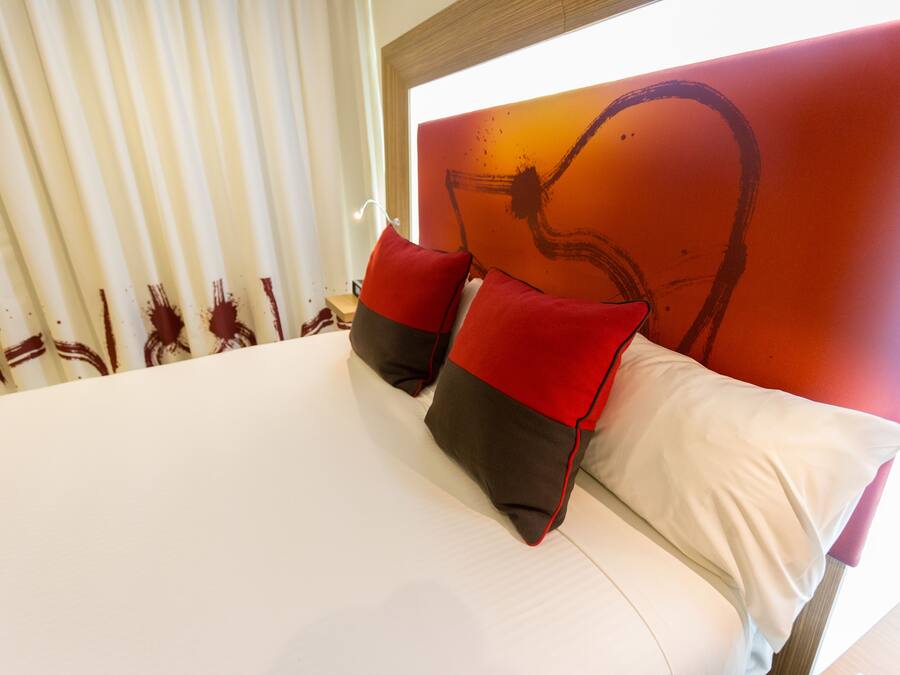 Quarto de hotel com uma cama grande, duas almofadas vermelhas e uma branca, cabeceira de madeira e uma janela com persianas.