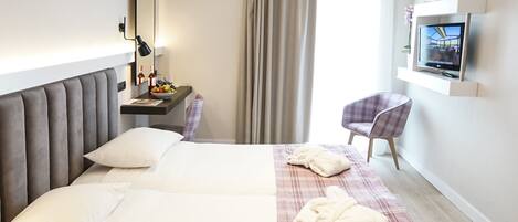 Quarto de hotel com uma cama grande, uma mesa de cabeceira com abajur, uma cadeira, uma TV e uma janela com cortinas.