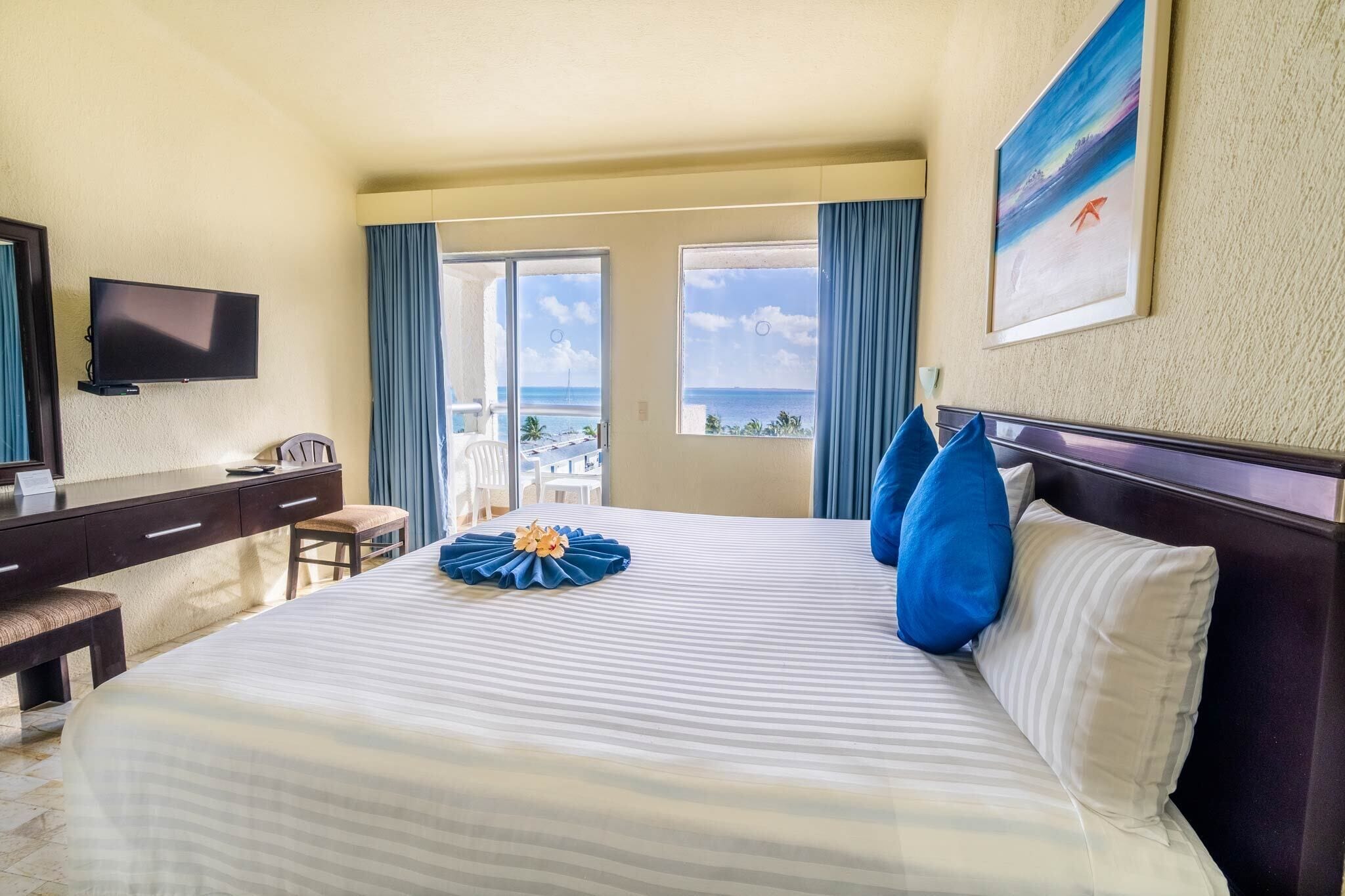 Quarto de hotel com uma cama grande, uma mesa, uma cadeira, uma televisão e uma varanda com vista para o mar.