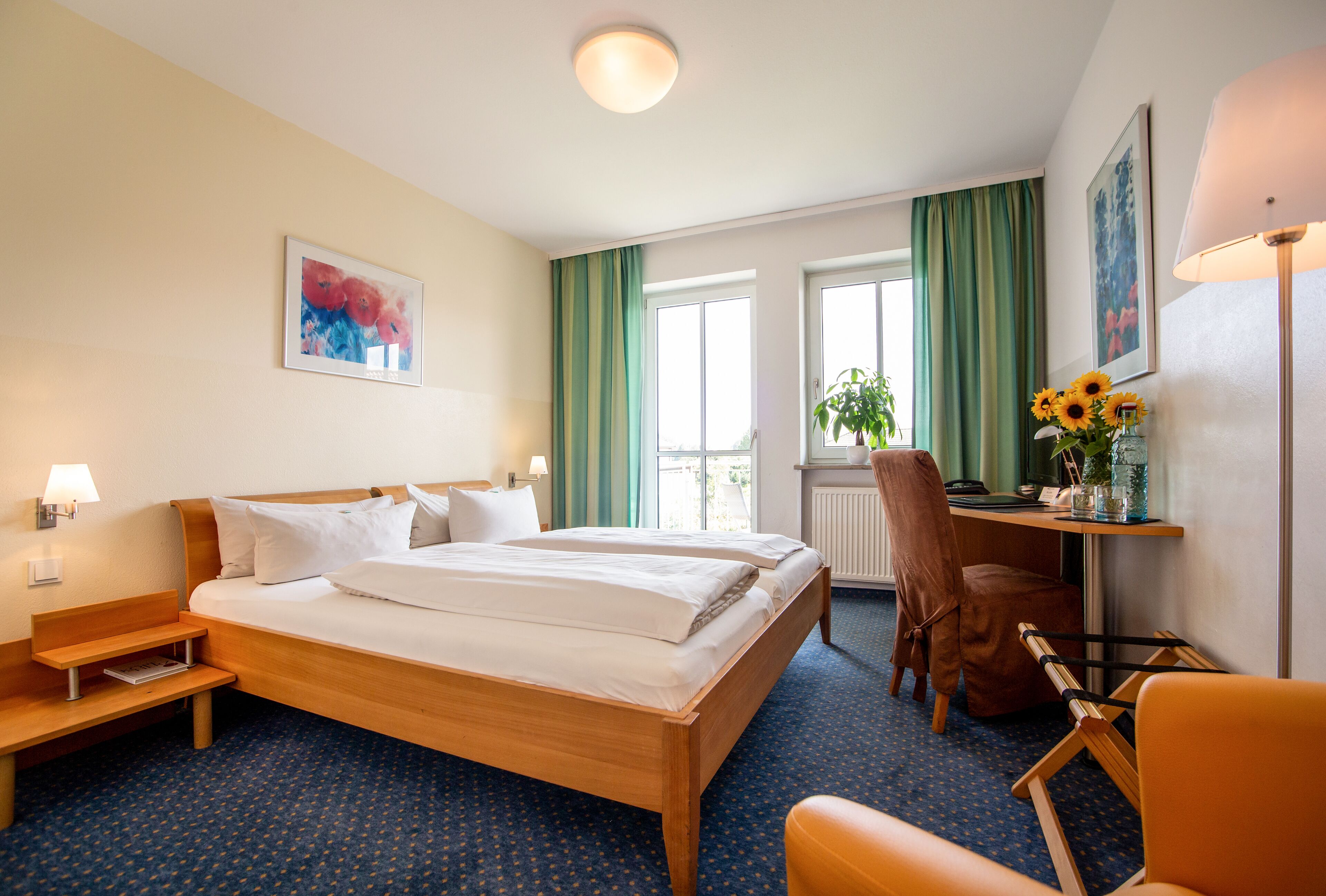 Quarto de hotel com uma cama grande, uma escrivaninha, uma cadeira, uma mesa de cabeceira e uma janela com cortinas.