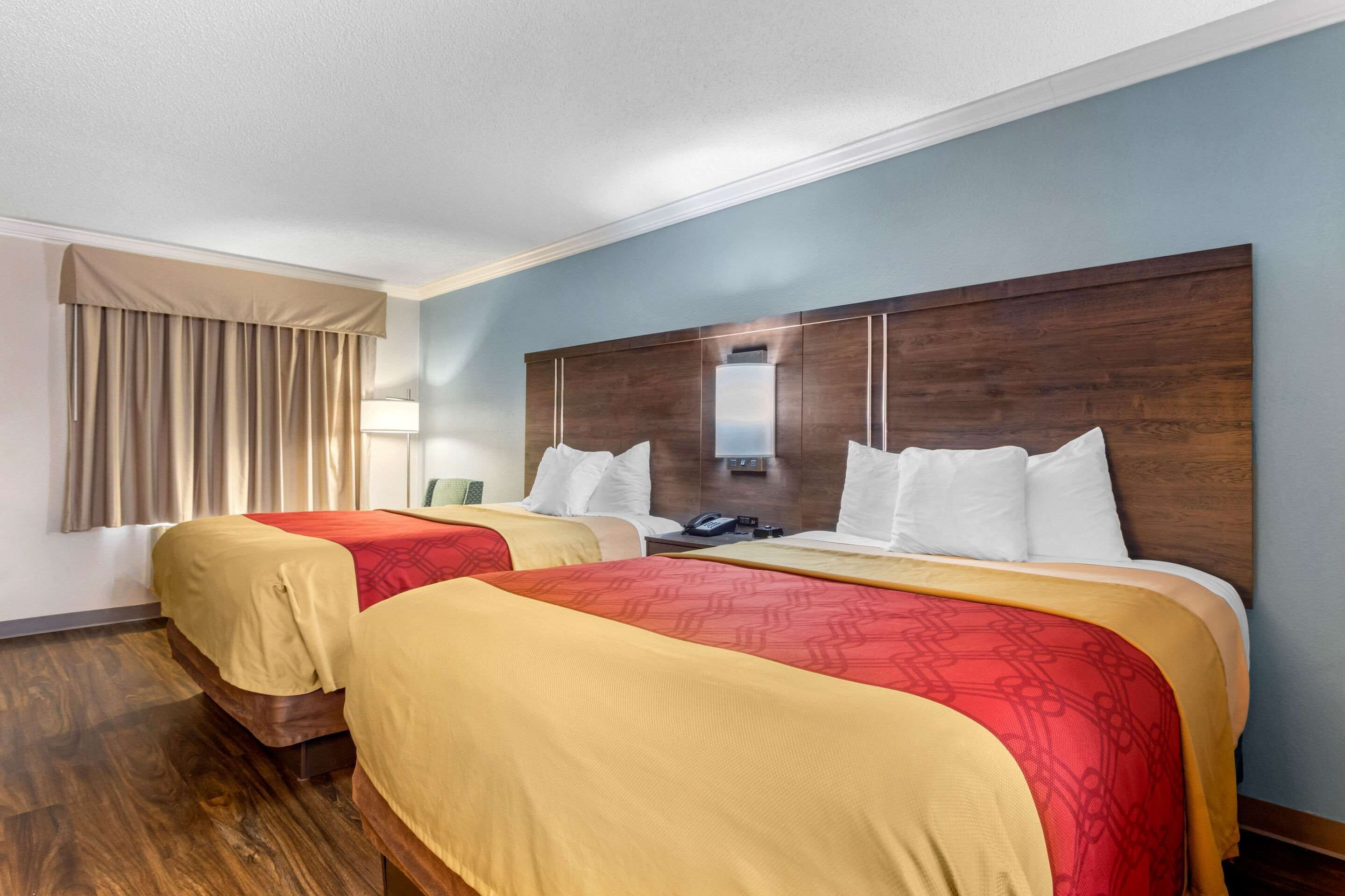 Quarto de hotel com duas camas, cabeceiras de madeira, uma janela grande com cortinas e uma mesa de cabeceira com abajur.