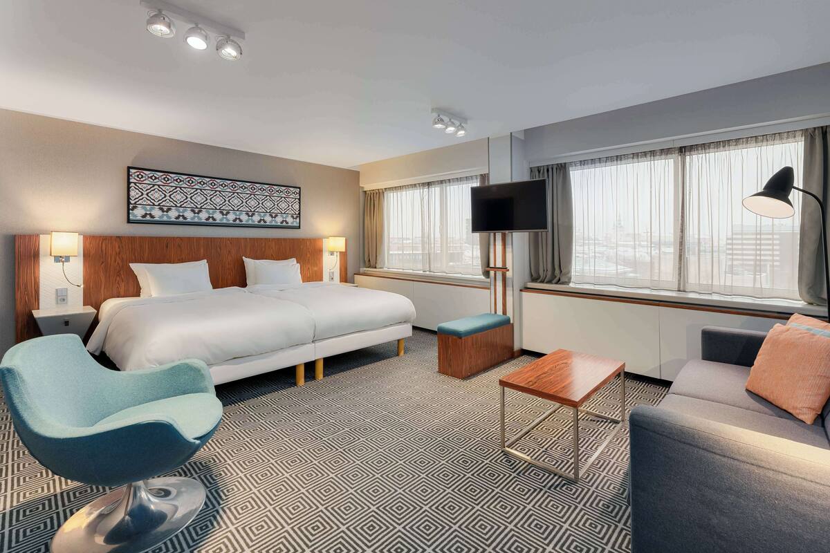 Quarto de hotel com cama, televisão, escrivaninha, cadeira e sofá.