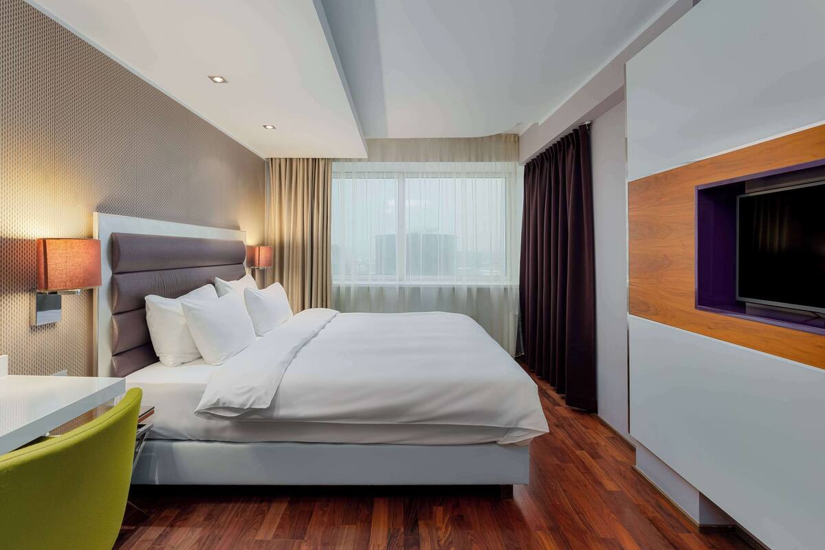 Quarto de hotel moderno com uma cama grande, uma escrivaninha com cadeira, televisão na parede e uma janela ampla com cortinas.