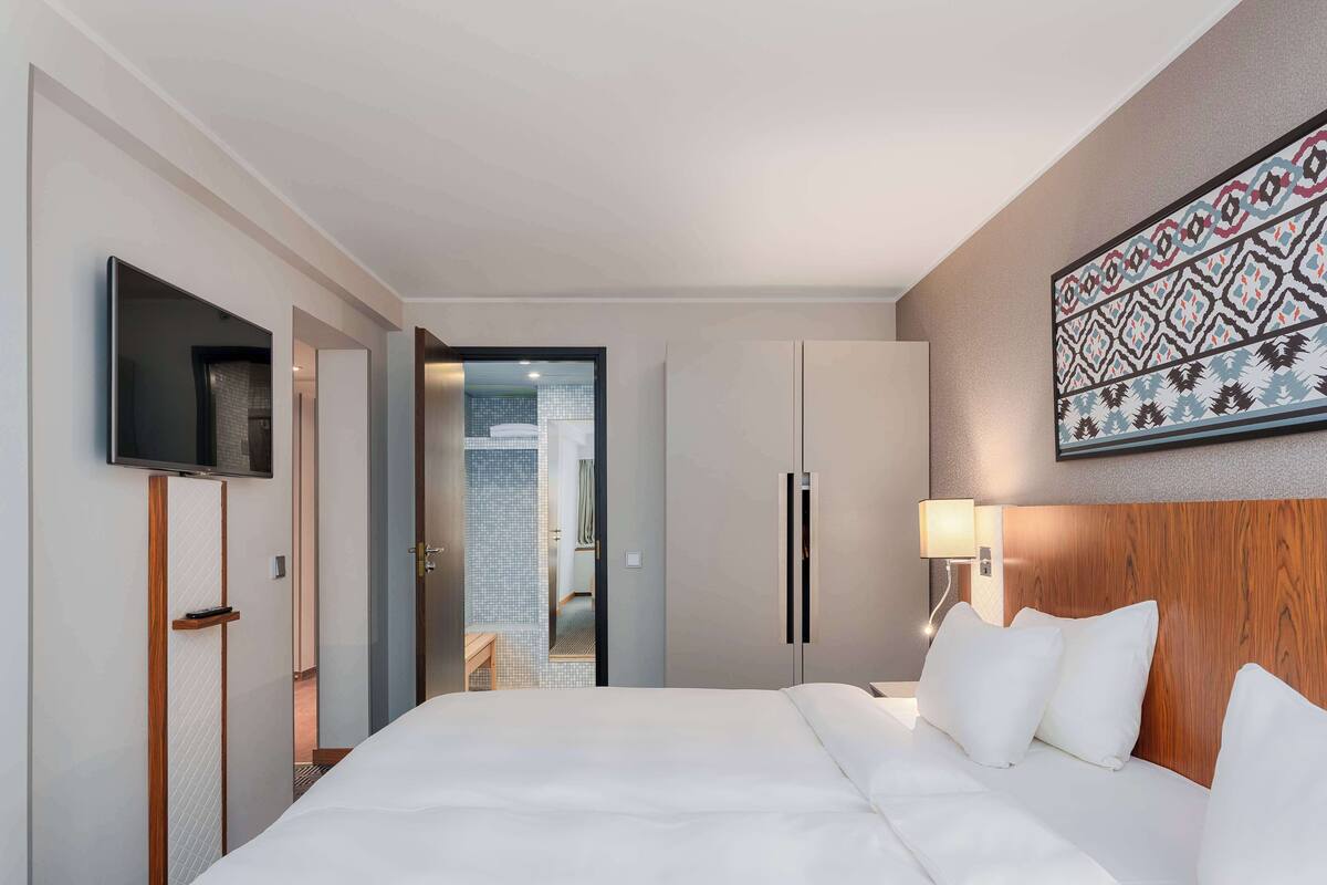 Quarto de hotel com uma cama grande, televisão fixada na parede, cabeceira de madeira, abajur e porta para o banheiro.