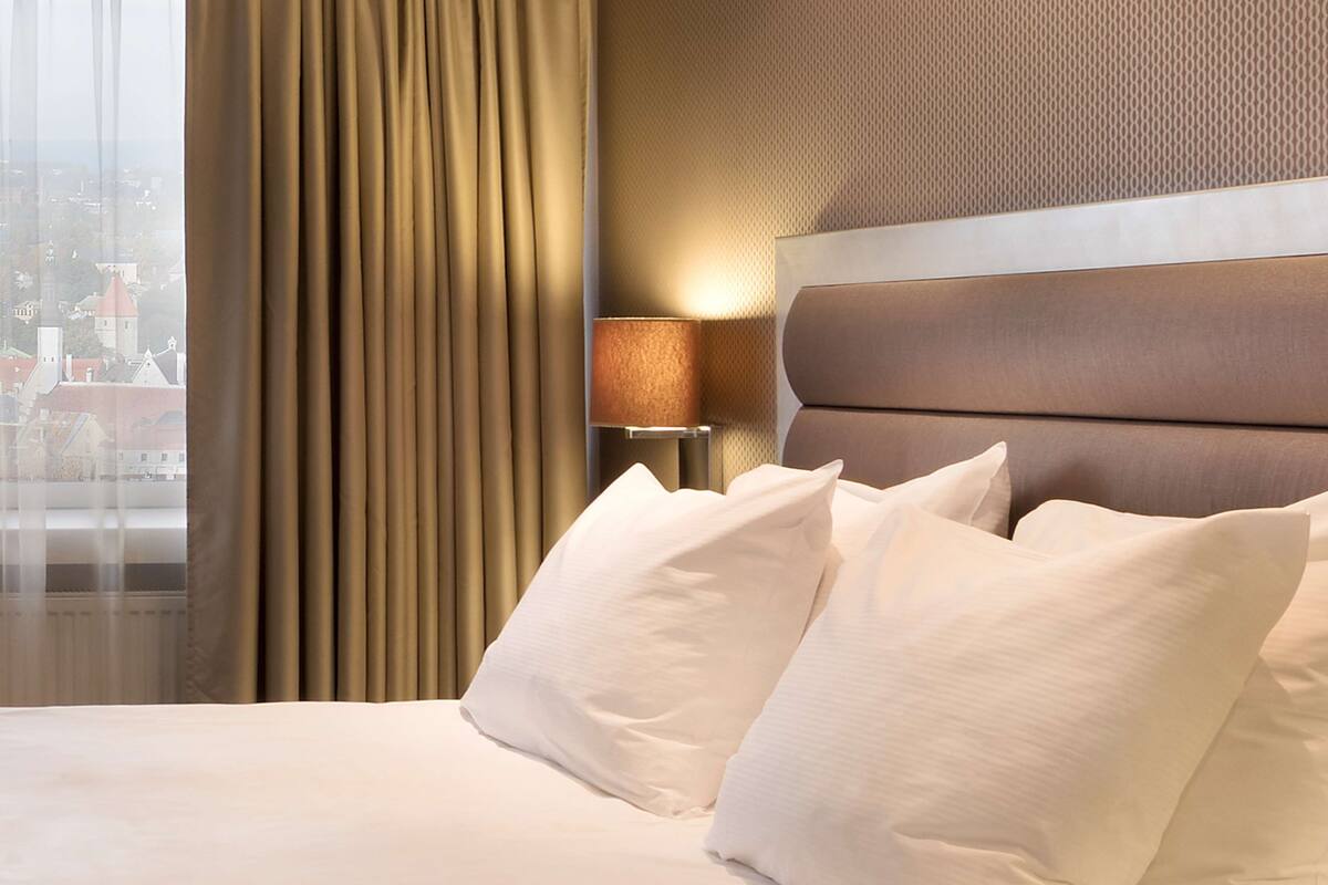 Quarto de hotel com uma cama, cortinas bege, uma luminária e vista para edifícios pela janela.