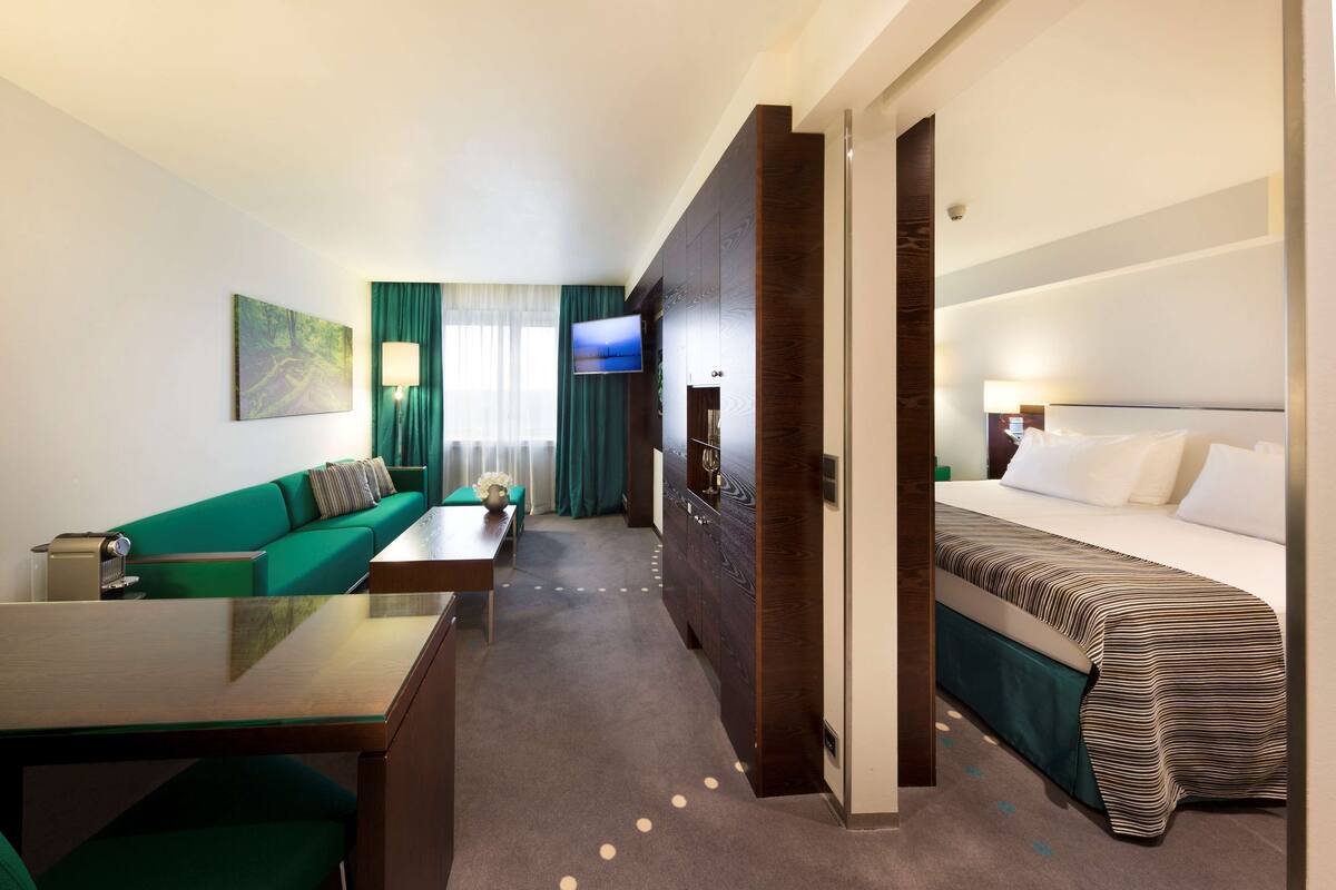 Quarto de hotel com sofá verde, cama com colcha listrada, televisão de tela plana na parede e escrivaninha com luminária.