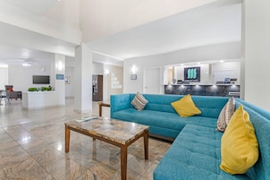 Uma sala de estar moderna com um sofá verde-água, mesa de centro de madeira e almofadas amarelas. No fundo, uma recepção com uma televisão e uma placa com a inscrição