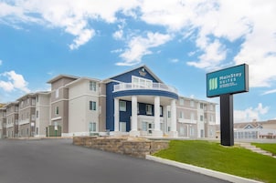 Um hotel MainStay Suites com fachada azul e branca, entrada central com colunas e uma placa de identificação.
