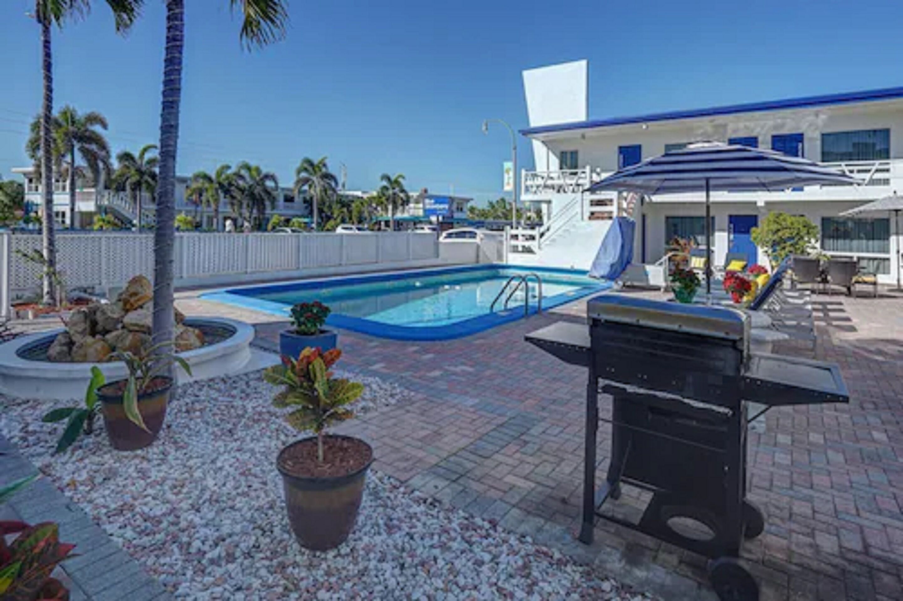 Uma área de piscina com piscina azul cristalina, palmeiras e um edifício com telhado azul e paredes brancas.