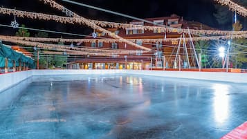 Pista de patinação no gelo em ambiente interno, com luzes pendentes e um edifício ao fundo.