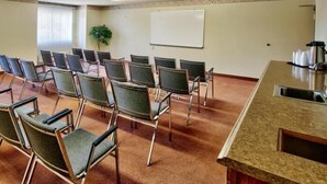 Sala de conferência com fileiras de cadeiras, um quadro branco e um espaço de cozinha.