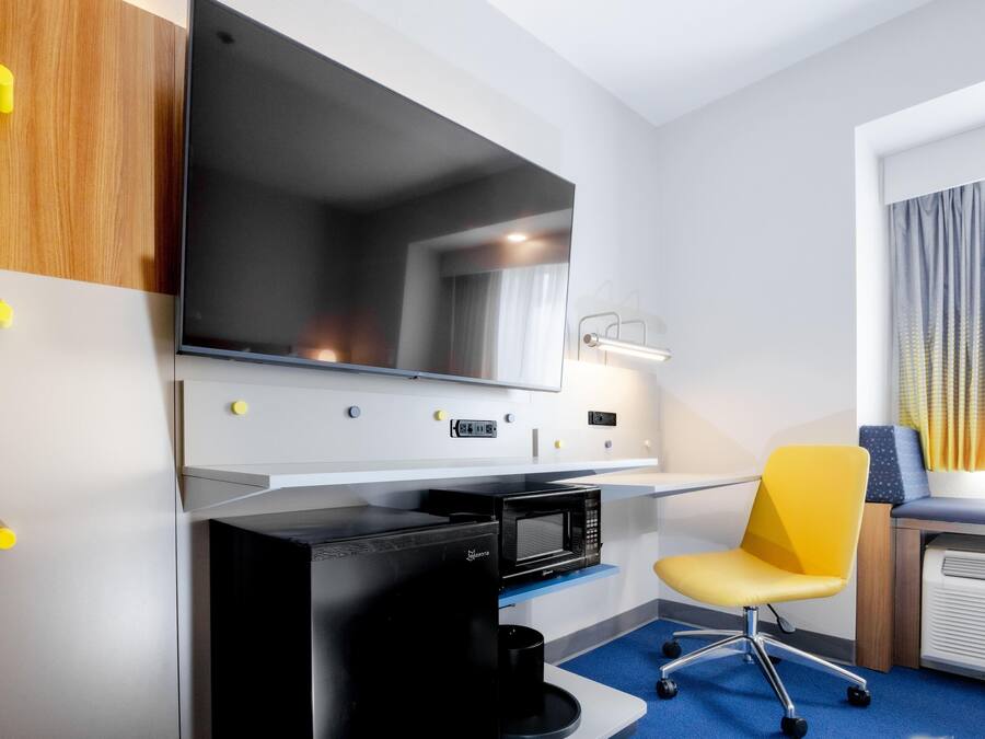 Quarto de hotel moderno com televisão de tela plana, cafeteira, uma cadeira amarela e escrivaninha.
