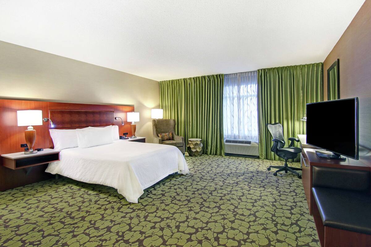Quarto de hotel com cama, duas mesas de cabeceira com luminárias, escrivaninha com televisão e uma cadeira.