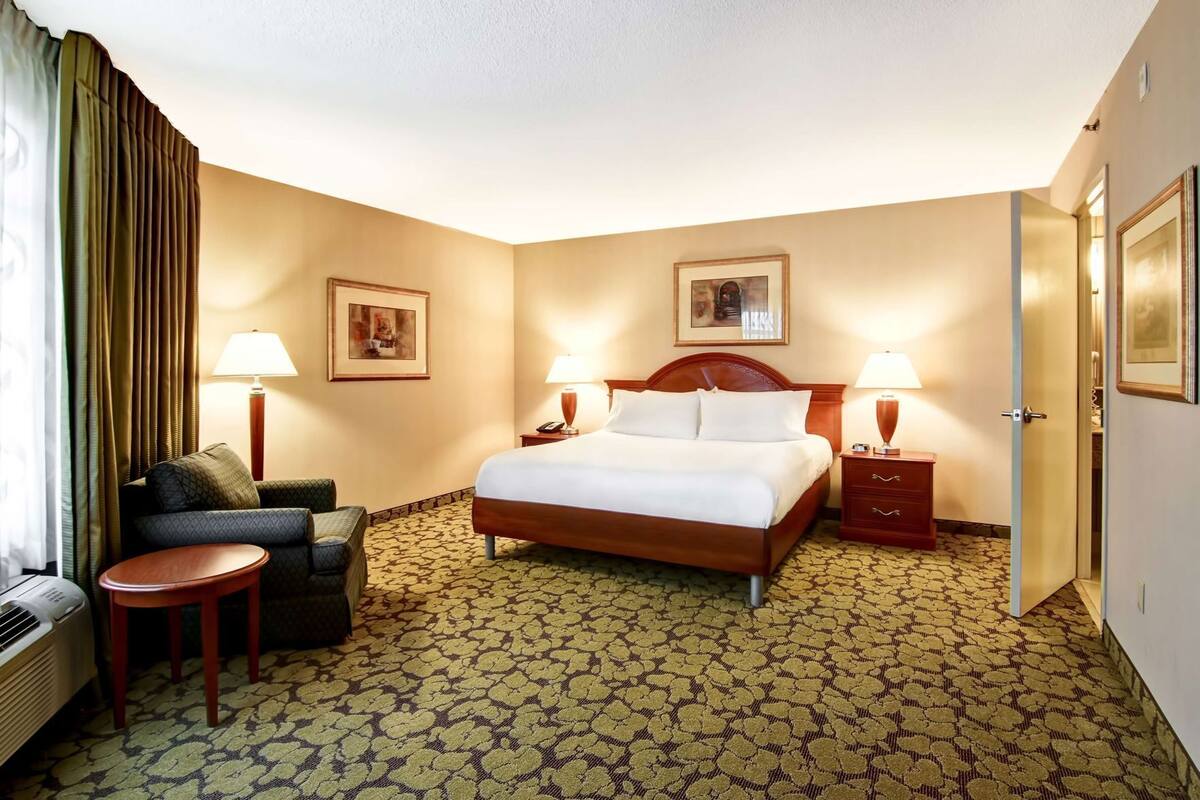 Quarto de hotel com cama, uma cadeira, uma mesinha, duas luminárias de cabeceira e uma porta.