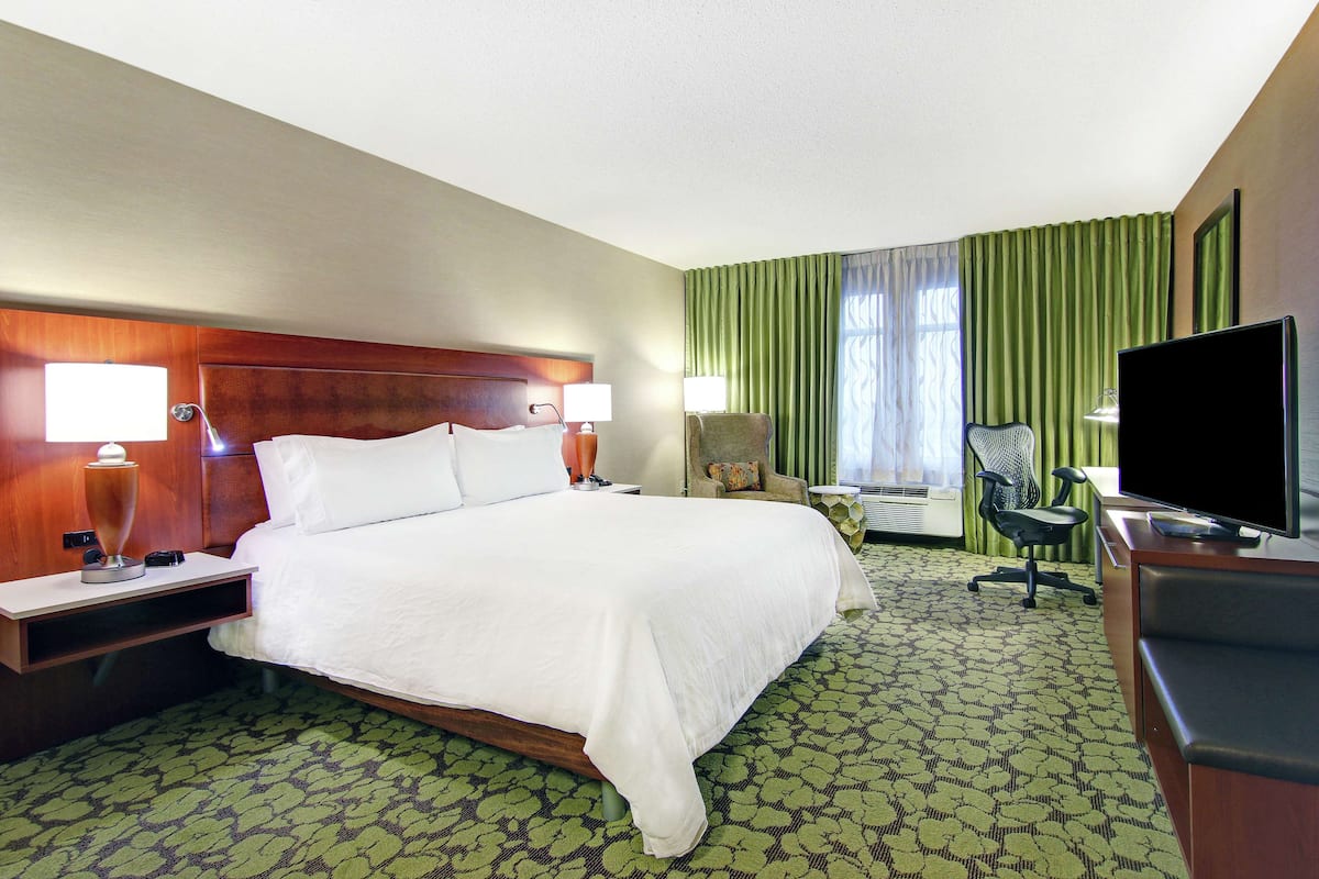 Quarto de hotel com uma cama grande, duas mesas de cabeceira com abajures, uma escrivaninha com televisão de tela plana, uma cadeira e uma janela grande com cortinas verdes.