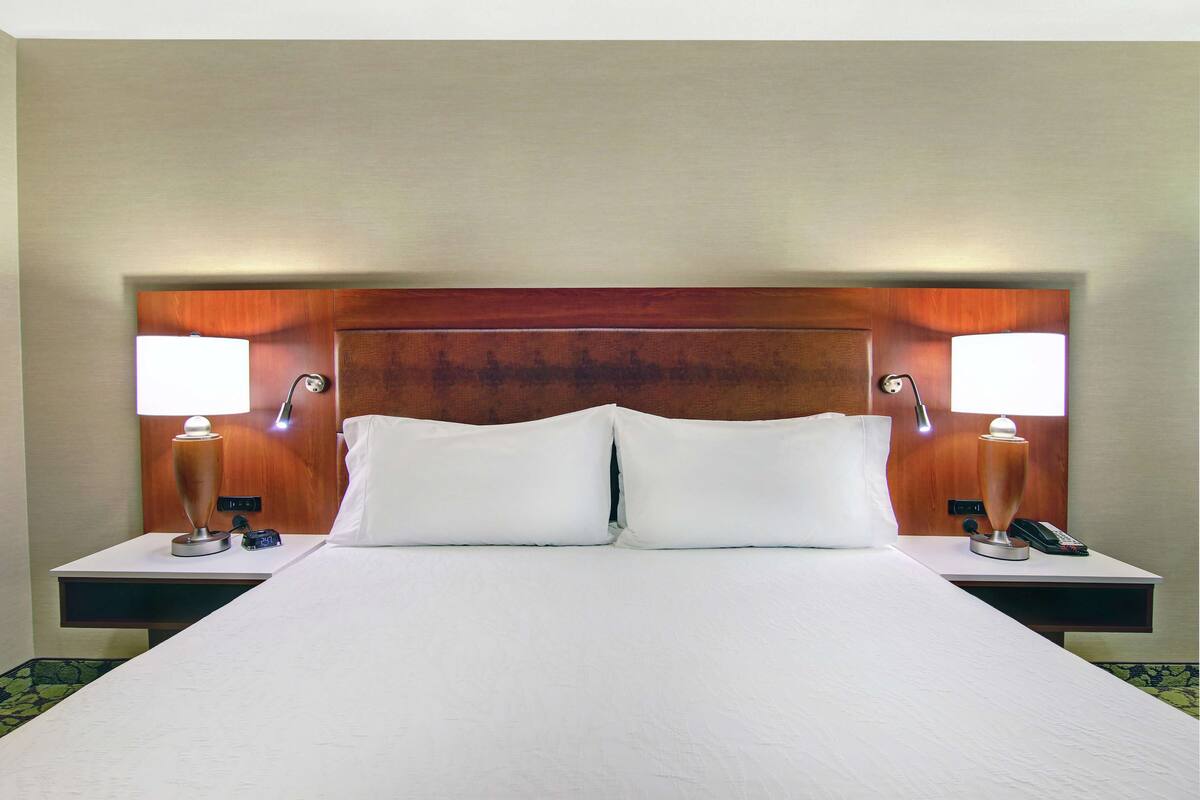 Quarto de hotel com uma cama grande, duas mesas de cabeceira com abajures e uma cabeceira de madeira.