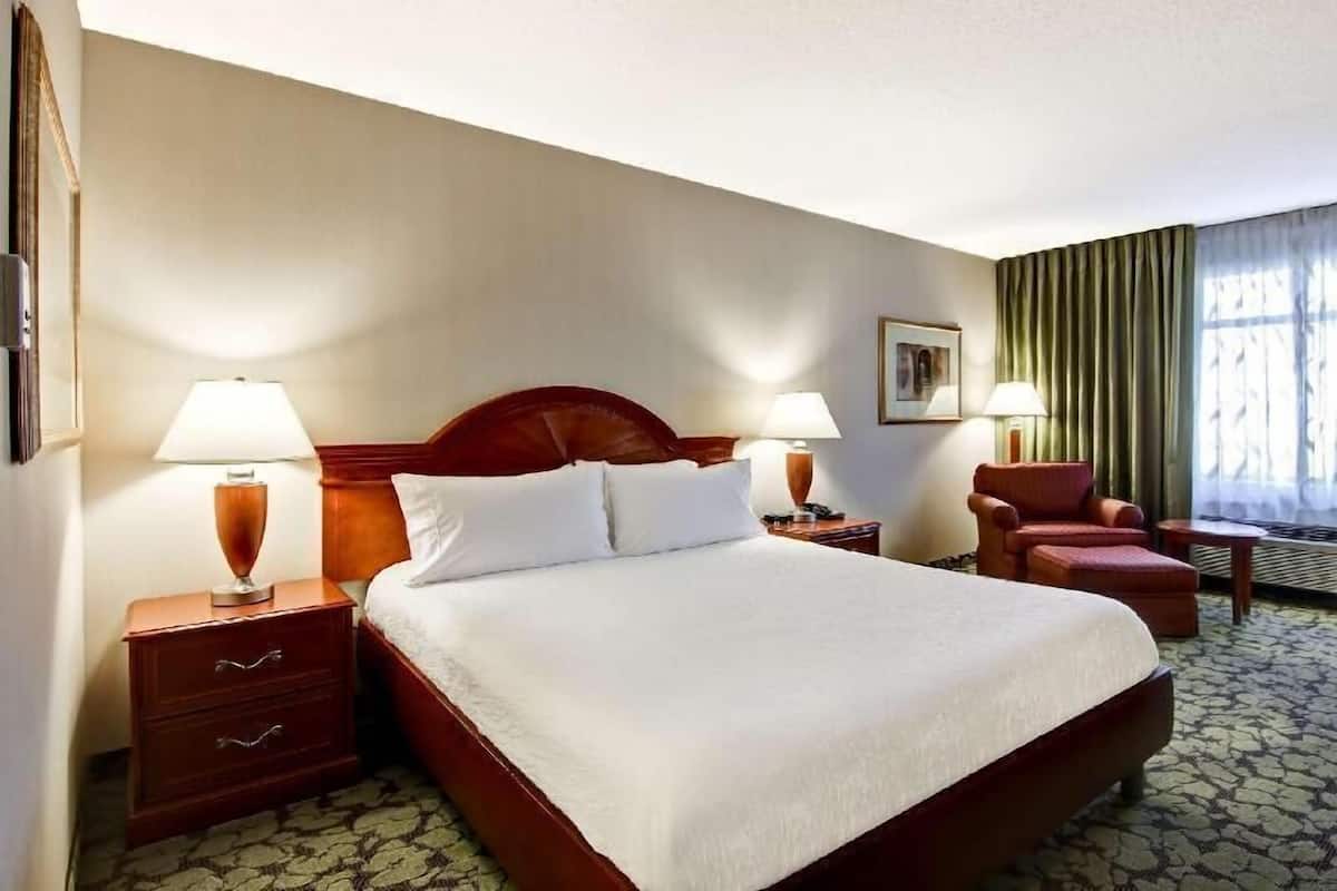 Quarto de hotel com uma cama grande, duas mesas de cabeceira com abajures, uma cadeira e uma janela com cortinas.
