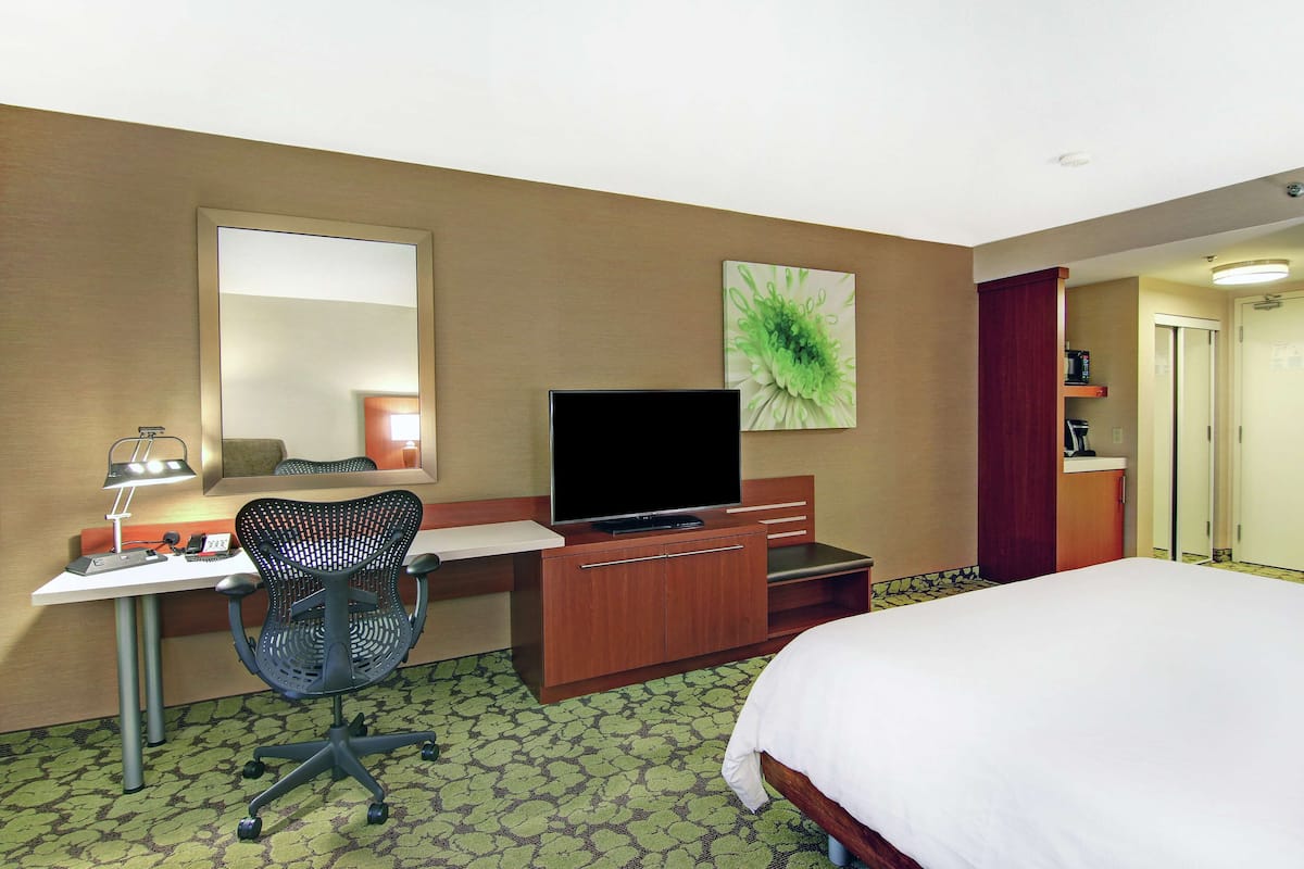 Quarto de hotel com cama, escrivaninha, cadeira, televisão e espelho.