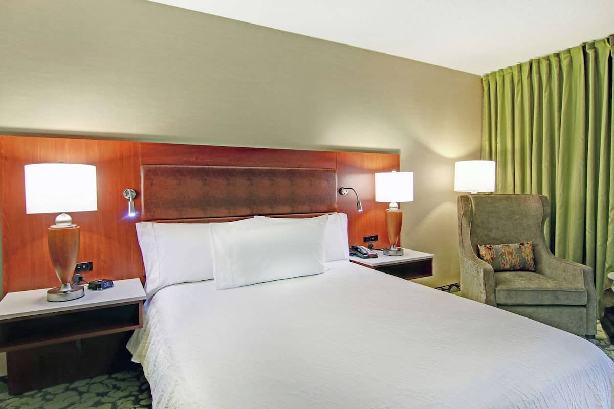 Quarto de hotel com uma cama grande, duas mesas de cabeceira com abajures, uma cadeira e uma cabeceira de madeira.