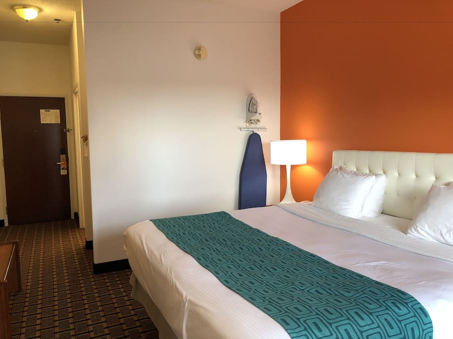 Quarto de hotel com cama, mesa de cabeceira, luminária, cabeceira e porta.
