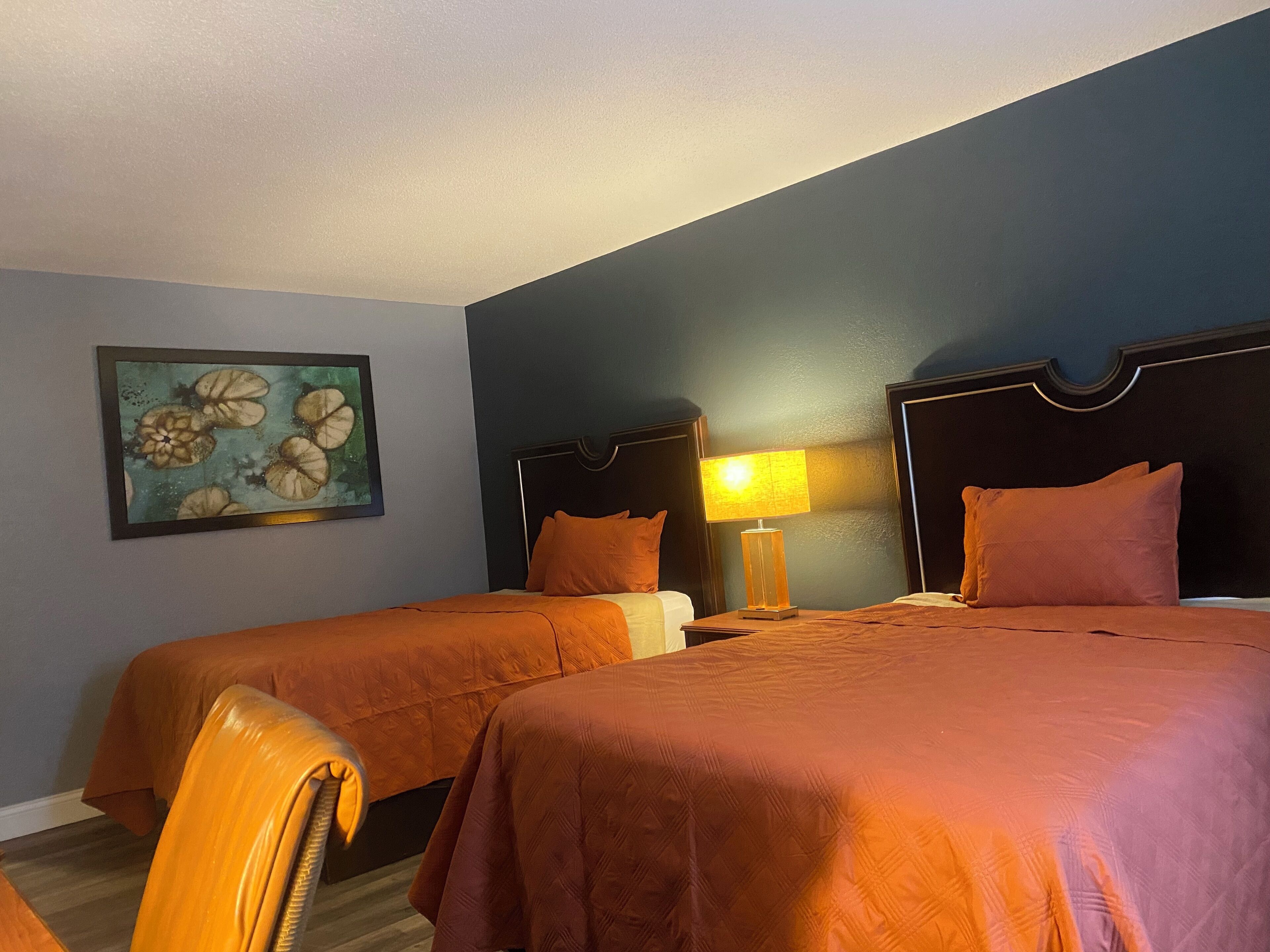 Quarto de hotel com duas camas, uma mesa de cabeceira com abajur e uma obra de arte emoldurada na parede.