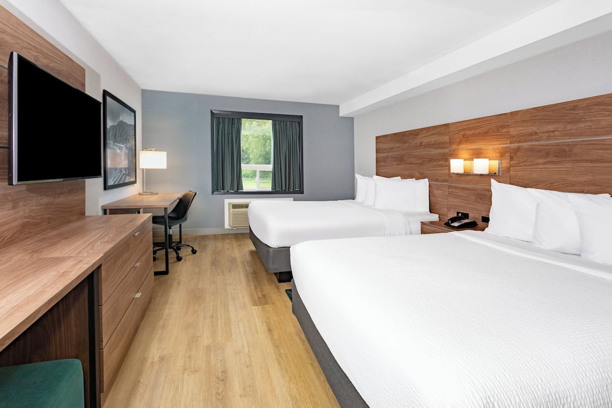Quarto de hotel com uma escrivaninha de madeira, duas camas, uma televisão, uma janela com cortinas e uma mesa de cabeceira com abajur.