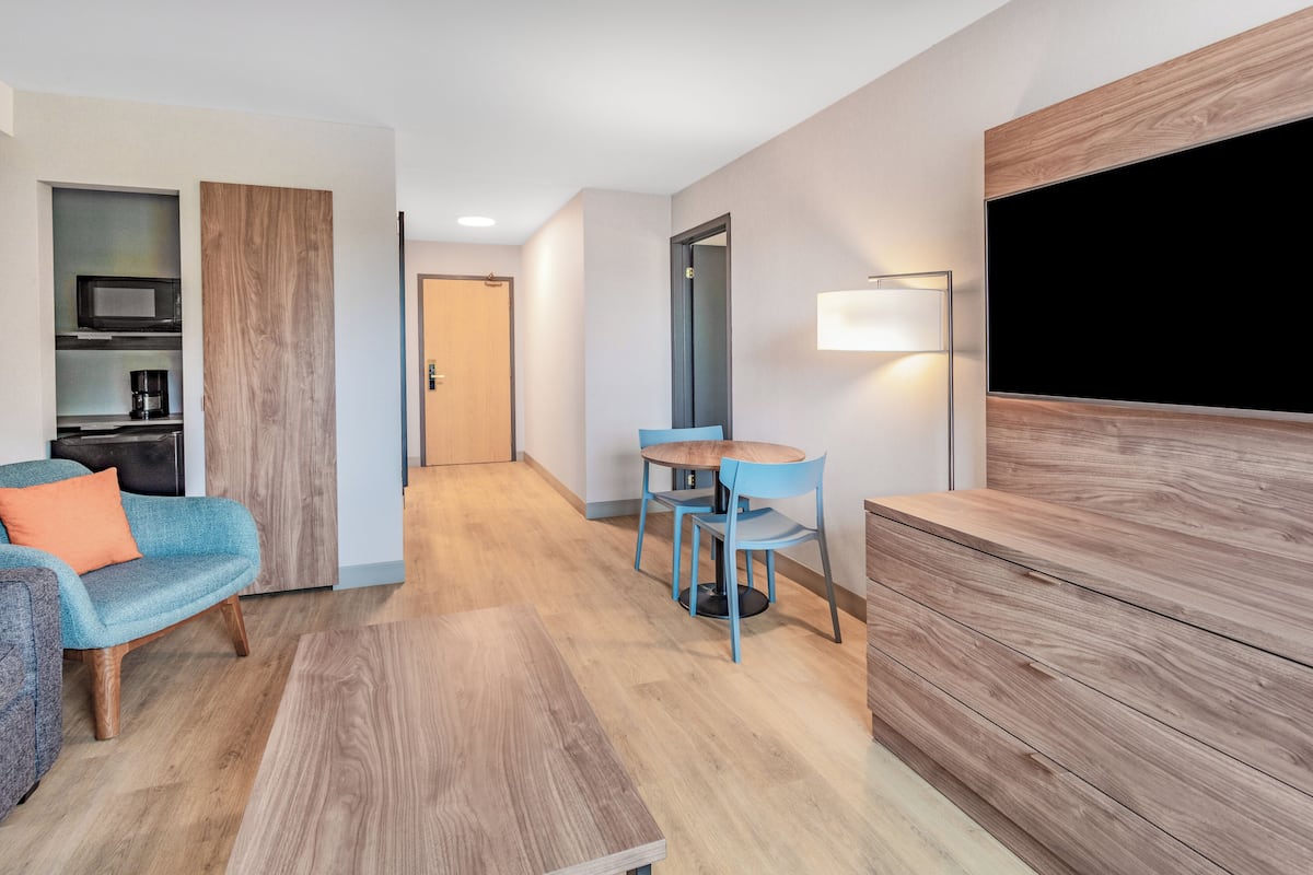 Quarto de hotel moderno com piso de madeira, um sofá, uma mesa de jantar com cadeiras, uma TV e um micro-ondas.