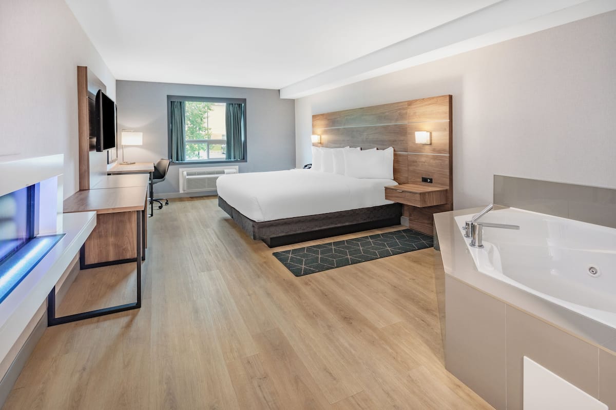 Quarto de hotel moderno com uma cama grande, uma escrivaninha com televisão de tela plana, luminária de mesa, janela com cortinas, televisão na parede, banheira integrada e um pequeno aquecedor.