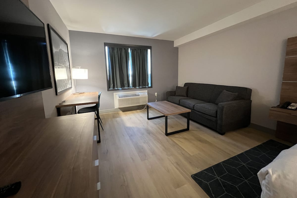 Quarto de hotel moderno com televisão de tela plana, sofá, mesa pequena e janela com cortinas.