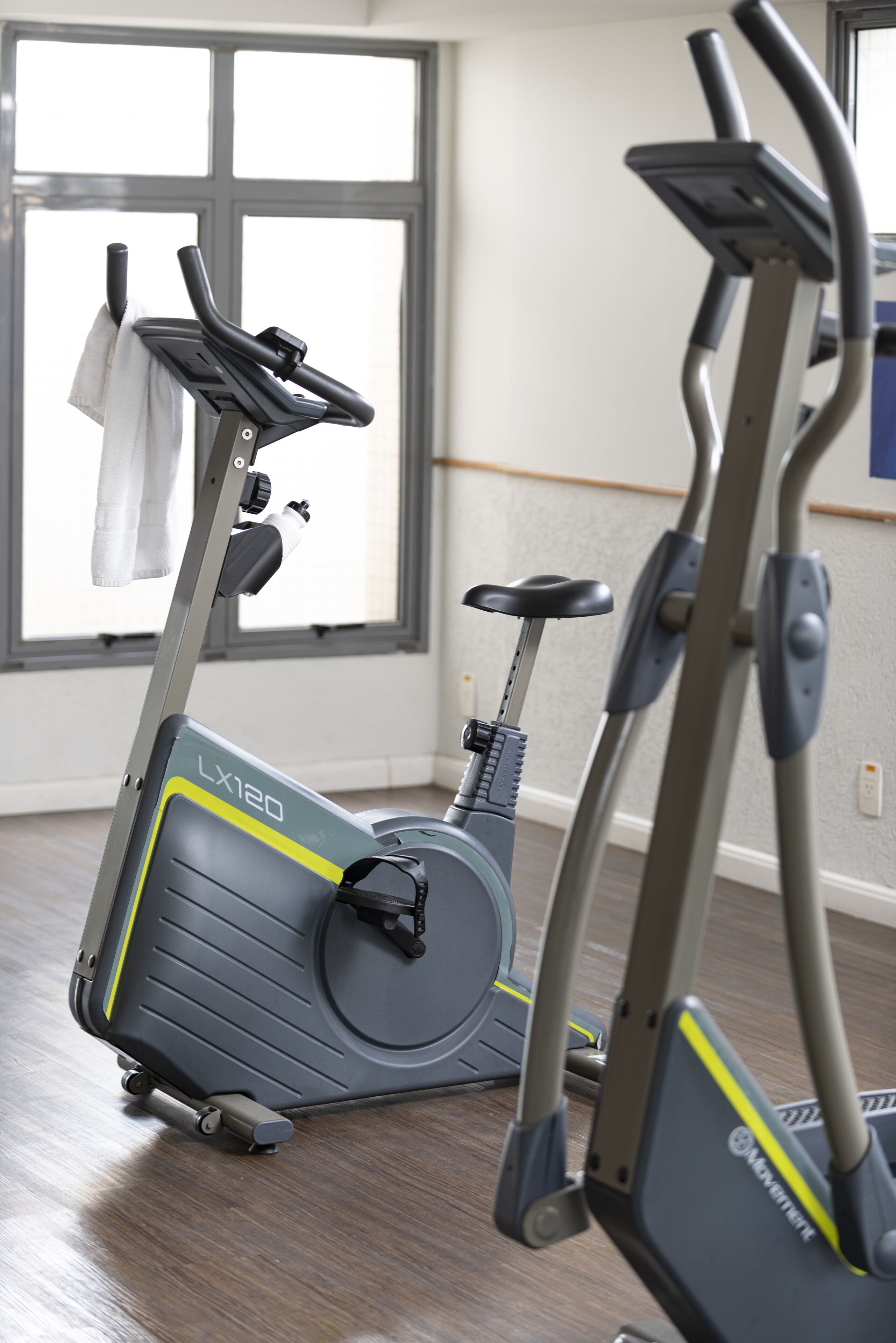 Uma máquina de exercícios com painel de controle e tela de exibição, localizada em um ambiente com uma janela grande e piso de madeira.