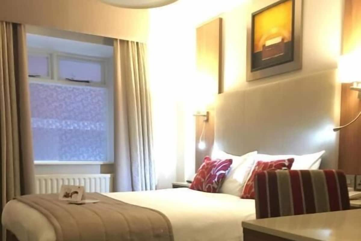 Quarto de hotel com cama, mesa de cabeceira, luminária, janela com cortinas e um quadro na parede.