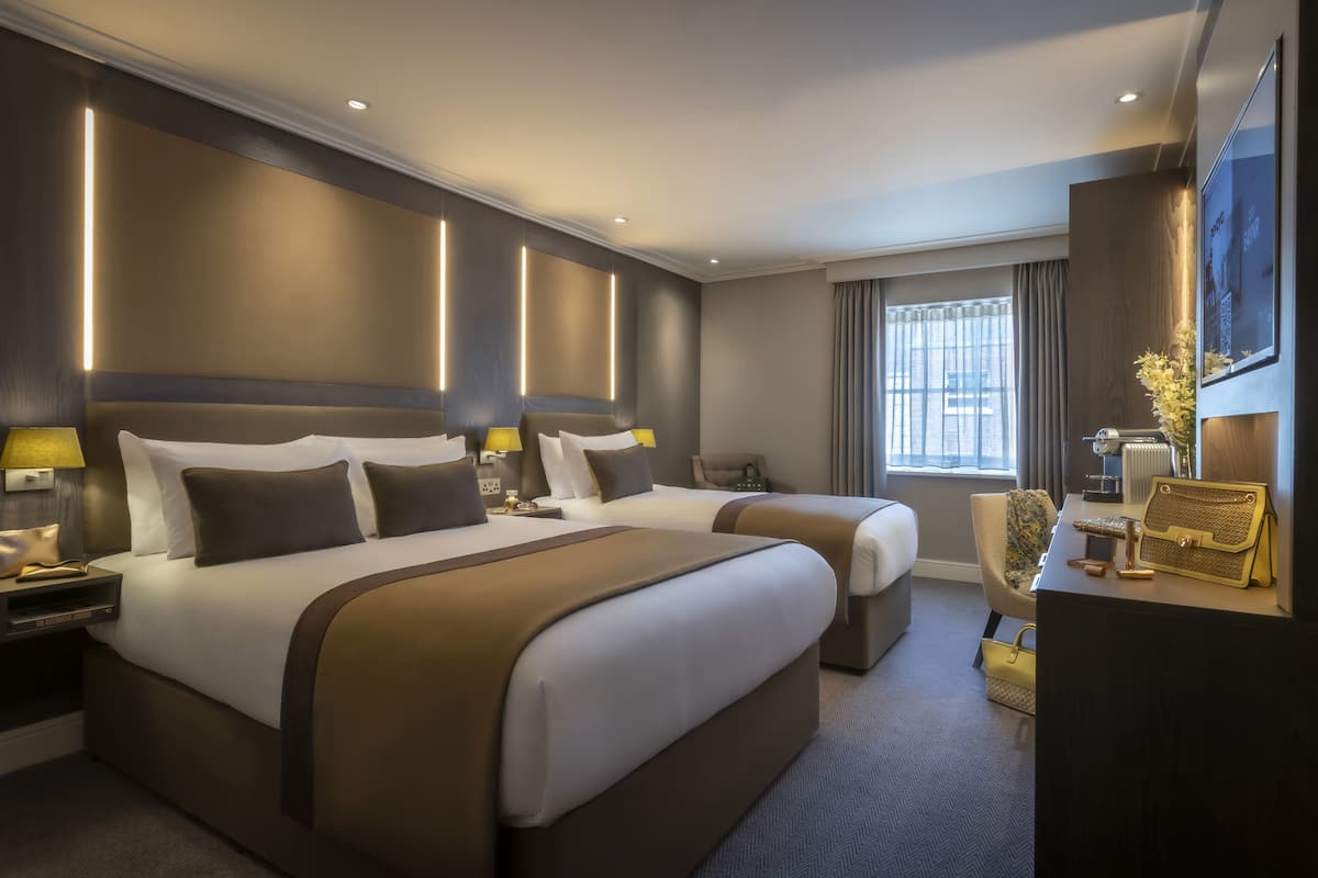 Quarto de hotel com duas camas, mesas de cabeceira com abajures, uma pequena escrivaninha com cadeira e uma janela grande com cortinas.