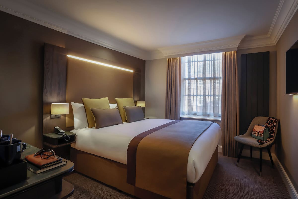 Quarto de hotel com uma cama grande, abajures, uma escrivaninha com um laptop e uma xícara, uma cadeira e uma televisão.