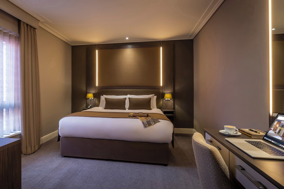 Quarto de hotel moderno com uma cama grande, abajures, uma mesa com um laptop e vista para a cidade pela janela.