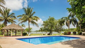 Piscina com um pavilhão de telhado de palha, cercada por palmeiras e vista para um lago.